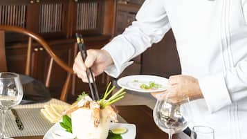 Um chefe de cozinha com uniforme branco e um chapéu de chef alto, preparando um prato sobre uma mesa com uma taça de vinho e um prato de comida.