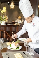 Um chefe de cozinha com uniforme branco e um chapéu de chef alto, preparando um prato sobre uma mesa com uma taça de vinho e um prato de comida.