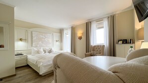 Quarto de hotel com uma cama grande, área de estar com uma poltrona, escrivaninha com luminária e janela com cortinas.