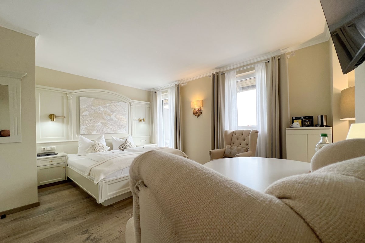 Quarto de hotel com uma cama grande, área de estar com uma poltrona, escrivaninha com luminária e janela com cortinas.