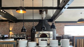 Interior de restaurante com parede de tijolos, painéis de madeira, luminárias pendentes, forno a pizza e área de bar com pratos empilhados.