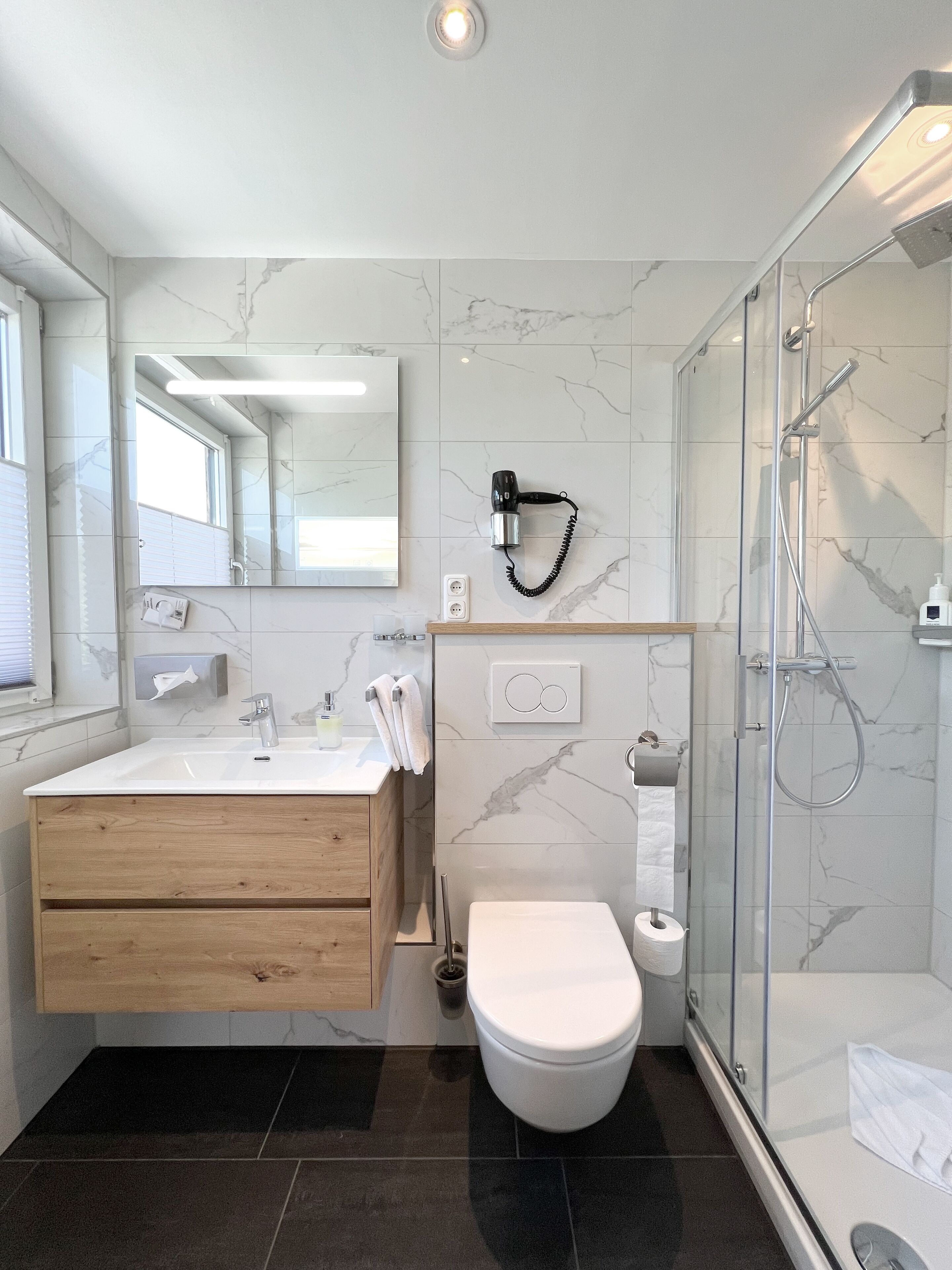Um banheiro moderno com painel de fundo de mármore, toucador de madeira, vaso sanitário e chuveiro com box de vidro.