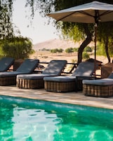 Uma área à beira da piscina com espreguiçadeiras e um guarda-sol, cercada por árvores e vista para uma paisagem montanhosa.