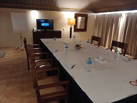 Uma sala de conferências com uma mesa comprida, cadeiras, garrafas de água, uma tela de projetor e uma televisão.