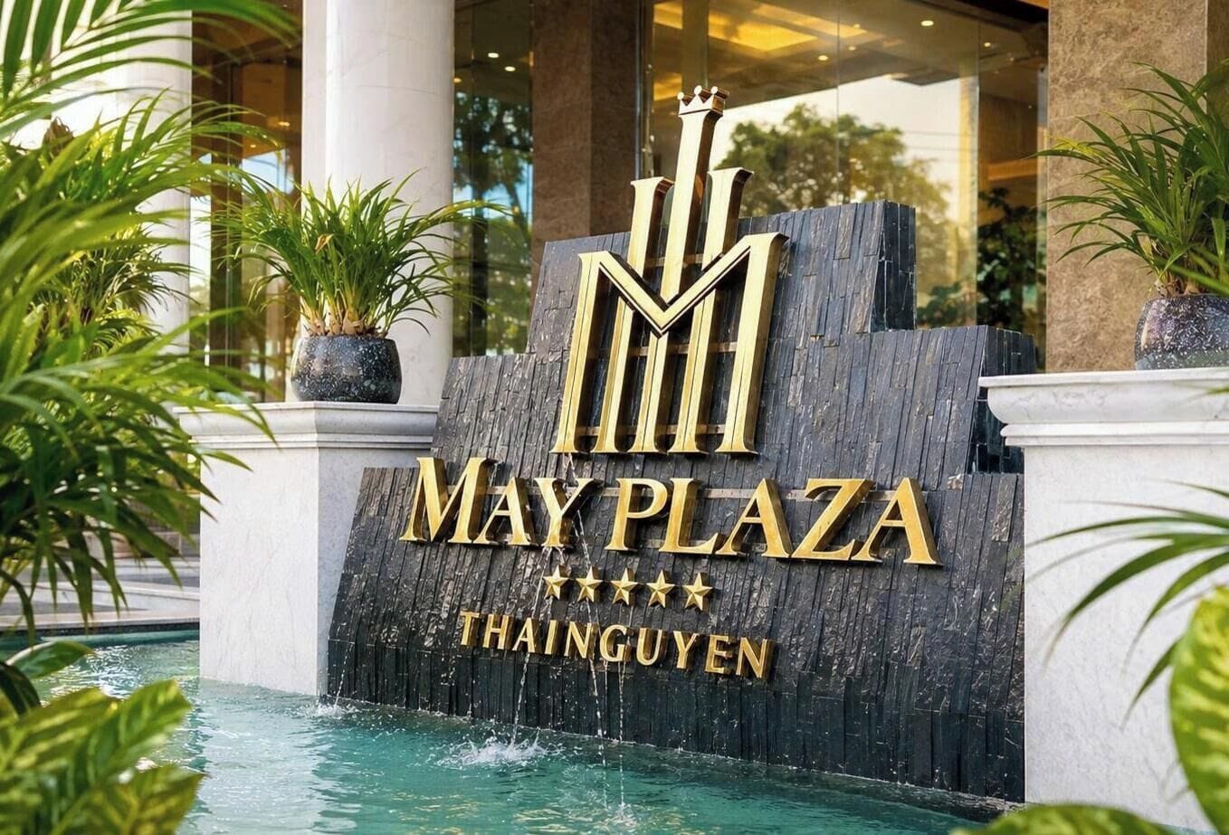 Entrada de hotel com uma placa indicando 'MAY PLAZA' e 'THAIGUYEN' em letras douradas, cercada por fontes e plantas em vasos.