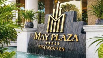 Entrada de hotel com uma placa indicando 'MAY PLAZA' e 'THAIGUYEN' em letras douradas, cercada por fontes e plantas em vasos.