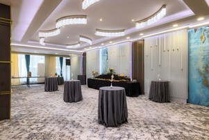 Ampla sala de conferências com pé direito alto, várias mesas com toques redondos e um buffet central, além de carpete estampado.