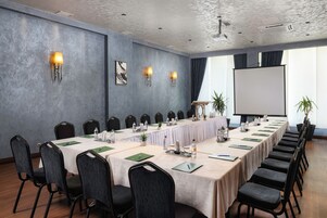 Sala de conferência com uma mesa comprida, cadeiras, tela de projetor e luminárias fixadas na parede.