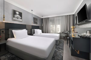 Quarto de hotel com duas camas, uma escrivaninha, televisão e janela com cortinas.