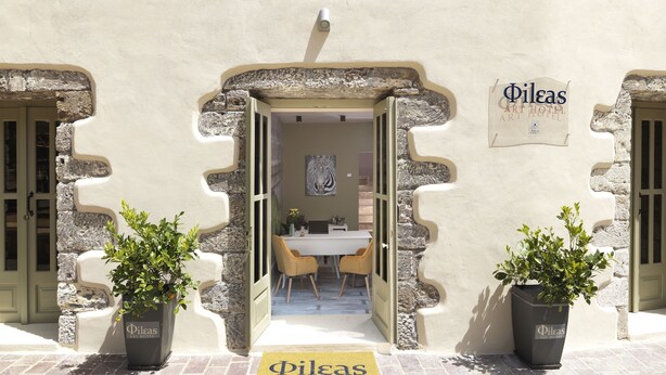 Entrada para o Pileas Art Hotel, com um arco de pedra, um balcão de recepção e duas plantas em vasos.