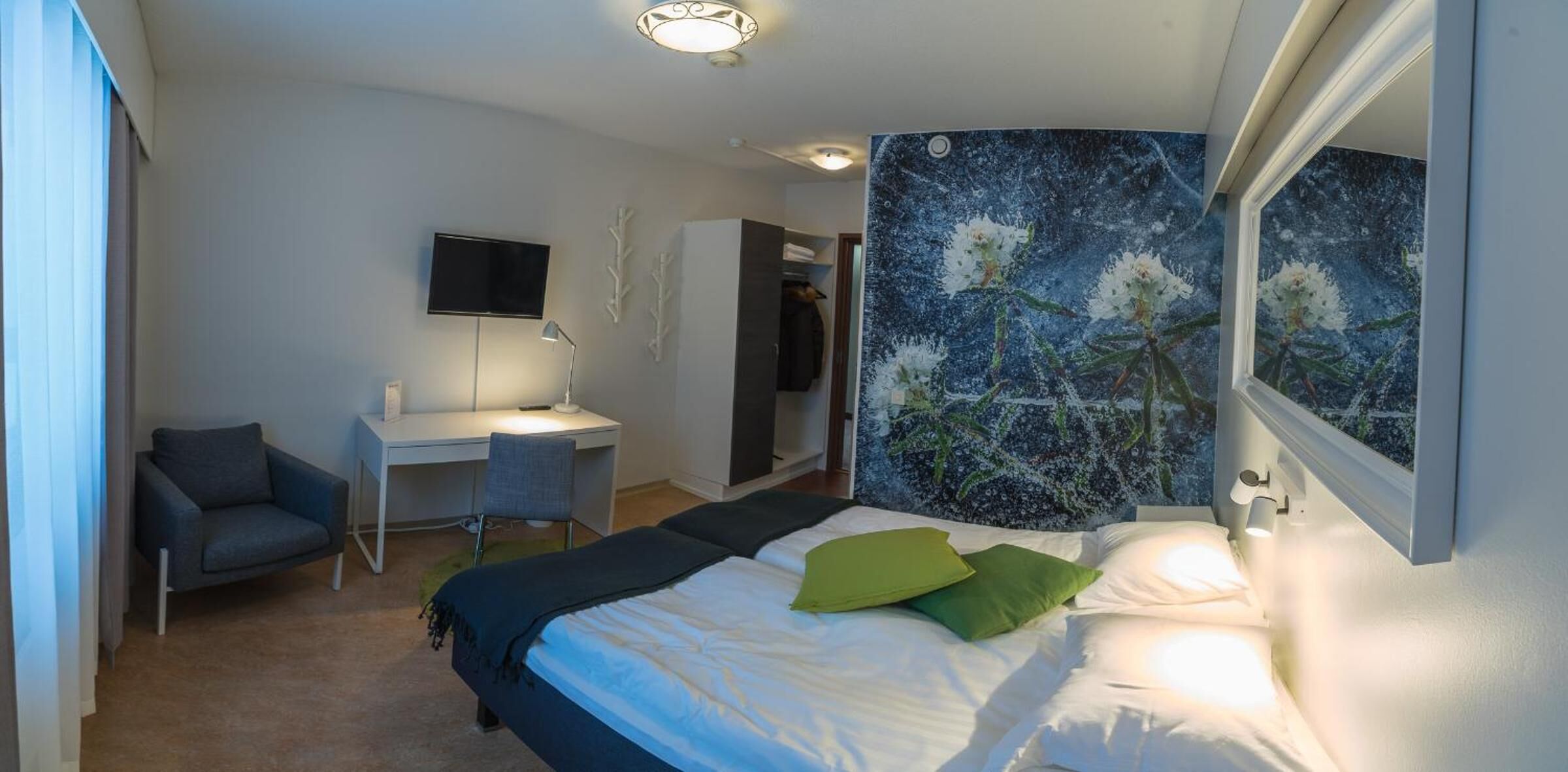Quarto com cama, escrivaninha, cadeira, televisão e parede com um mural floral.