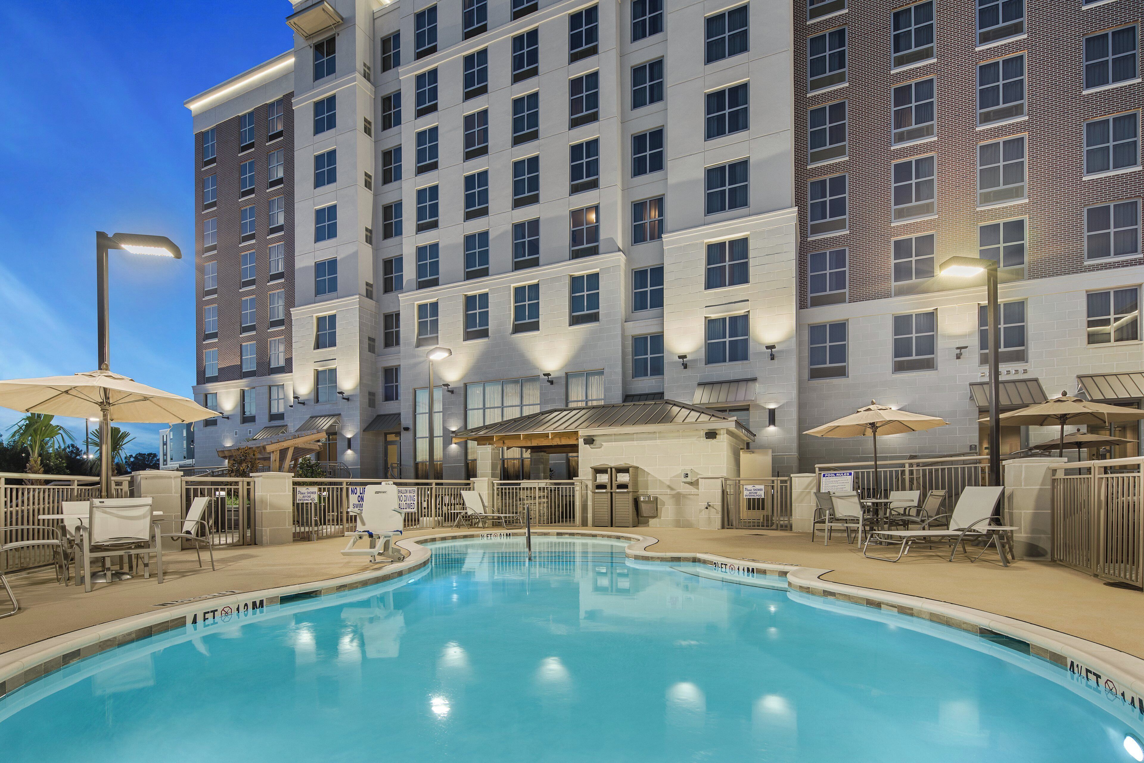 Uma área de piscina de hotel com piscina azul cristalina, espreguiçadeiras, guarda-sóis e um edifício com várias janelas ao fundo.