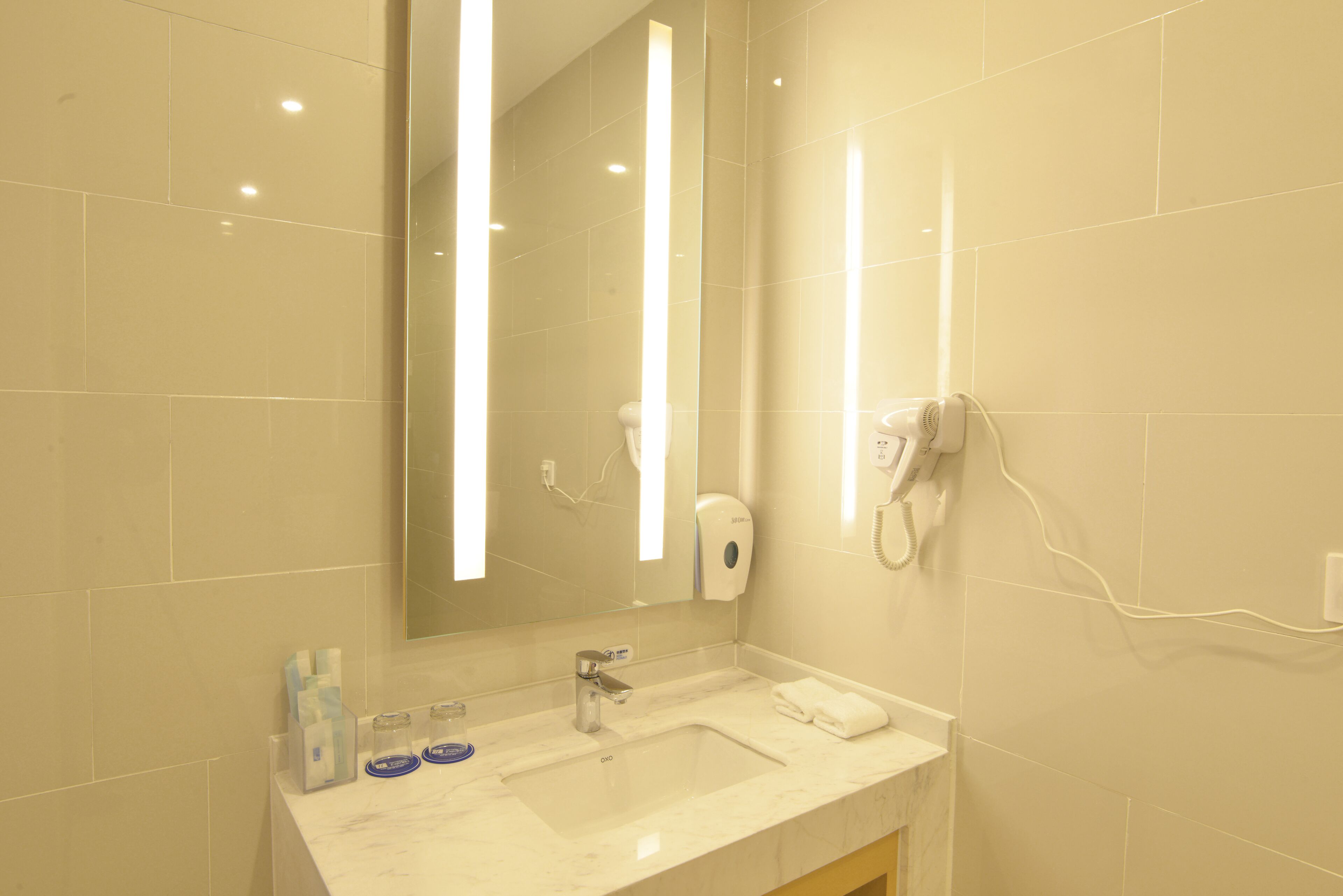 Casa de banho de hotel com um espelho grande, uma pia, um secador de cabelo e um dispensador de sabonete.