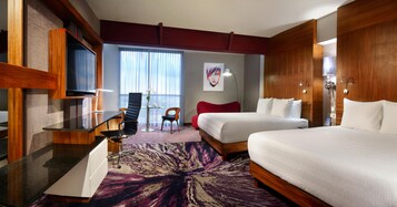 Quarto de hotel com cama, escrivaninha, cadeira, televisão e uma janela grande.
