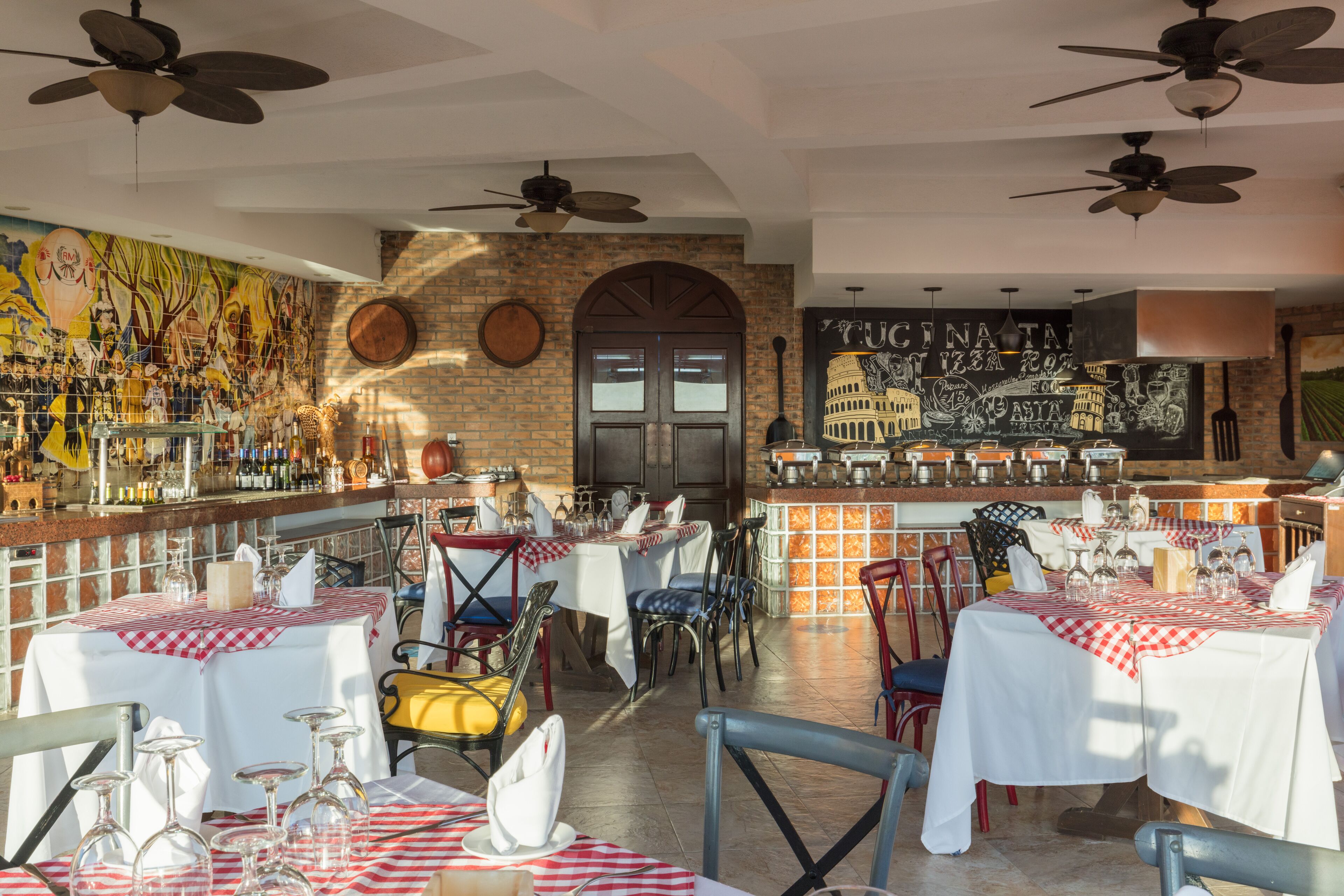 Um restaurante com várias mesas preparadas para refeições, um bar com diversos tipos de garrafas e um mural na parede.