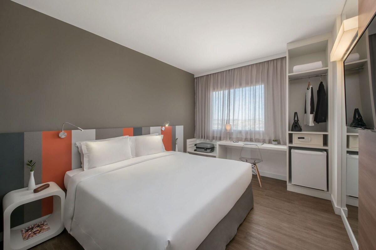 Quarto de hotel moderno com uma cama grande, uma escrivaninha, um guarda-roupa e uma janela com cortinas.