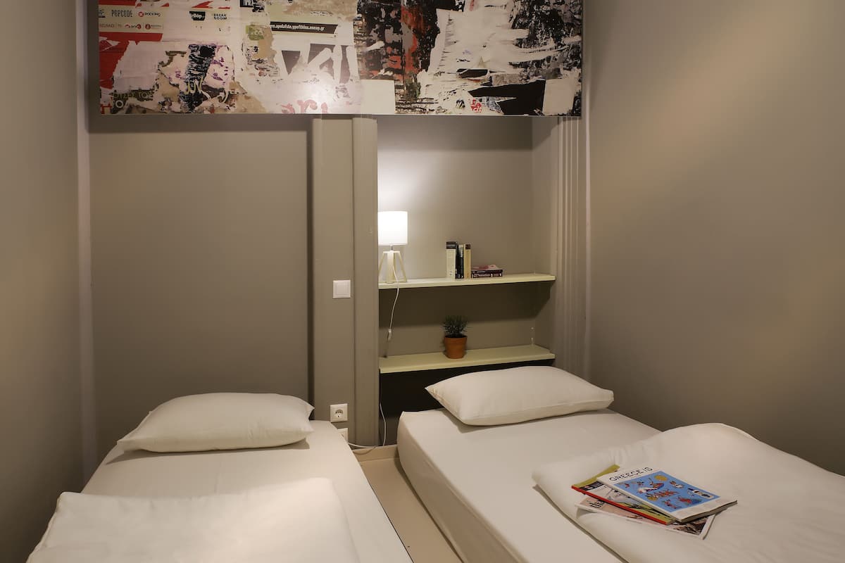 Quarto pequeno e limpo, com duas camas individuais, uma mesa de cabeceira com luminária, uma estante com livros e um painel decorado com posters.