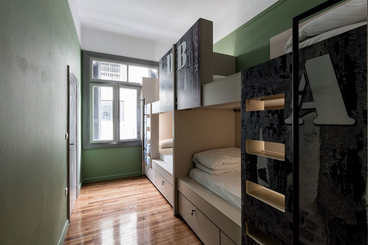 Quarto com beliches, janela com persianas e piso de madeira.