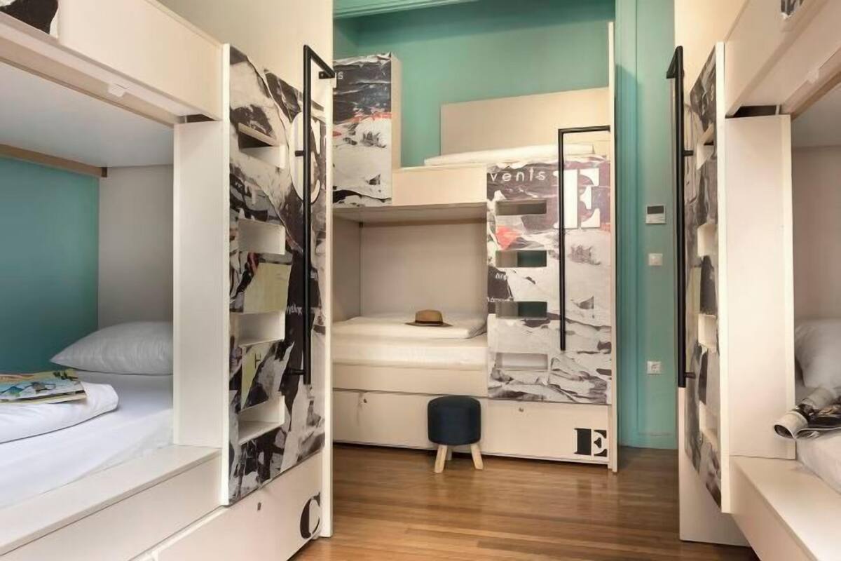 Quarto pequeno com beliches, um banquinho e prateleira na parede com vários objetos.