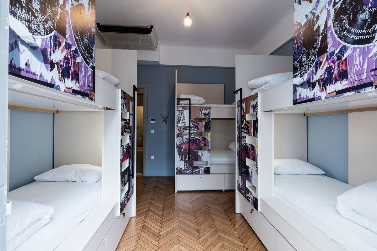 Um quarto de dormitório com beliches, piso de madeira em padrão herringbone e uma porta que dá para outro quarto.