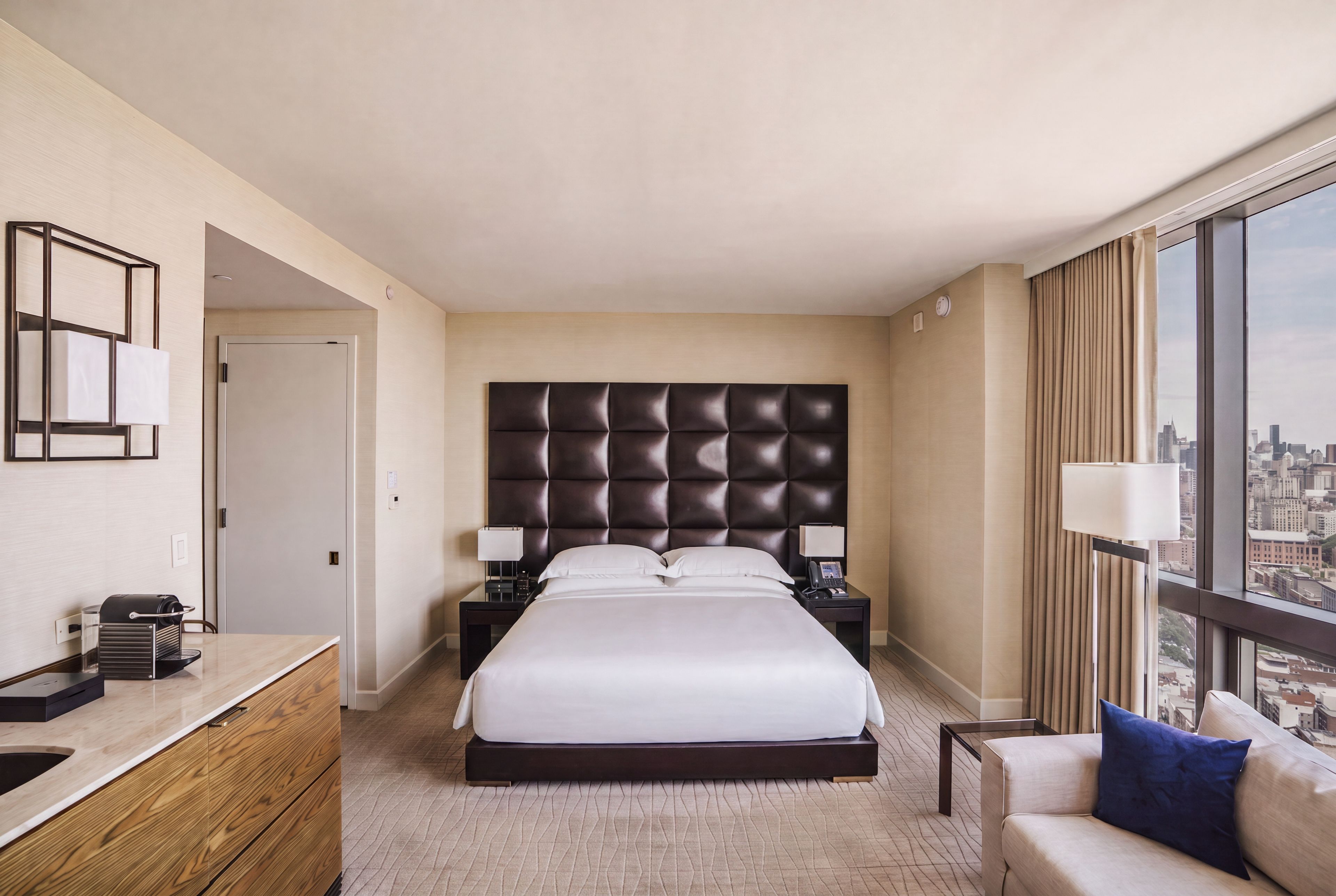 Quarto de hotel com uma cama grande, uma escrivaninha, uma cadeira, uma mesa de cabeceira, uma luminária e vista para a cidade.