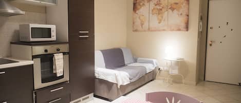Quarto de hotel compacto com cozinha compacta, cama com manta cinza, pequena mesa de cabeceira com abajur e uma pintura de mapa-múndi na parede.