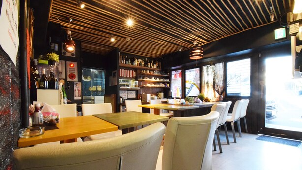 Interior de restaurante com mesas e cadeiras de madeira, prateleiras com garrafas e copos, e vista pela janela.