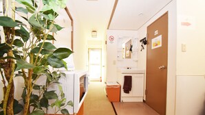 Corredor de hotel com balcão de recepção, uma planta, banheiro com pia e toalha, e uma porta com placa.