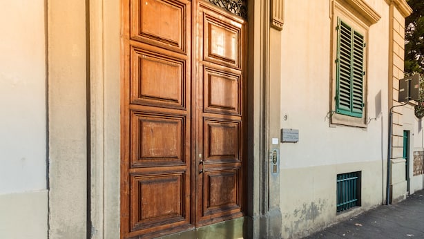 Uma grande porta dupla de madeira com detalhes em bronze, embutida em um edifício com fachada clara e uma pequena janela com persianas verdes.