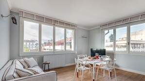 Sala de estar moderna com sofá, mesa de jantar e cadeiras. Há uma janela grande com vista para prédios.
