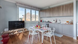 Cozinha moderna com armários brancos, mesa de jantar com cadeiras, televisão e janela com vista para a cidade.
