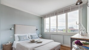 Quarto com uma cama grande, mesa de cabeceira com abajur, janela com persianas e vista da cidade.