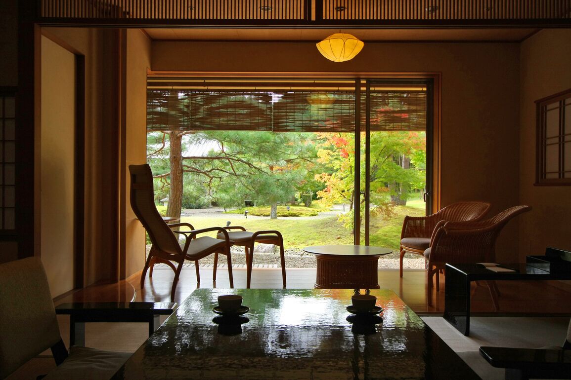 Quarto com uma mesa de jantar, cadeiras, uma luminária e vista para um jardim através de grandes janelas.