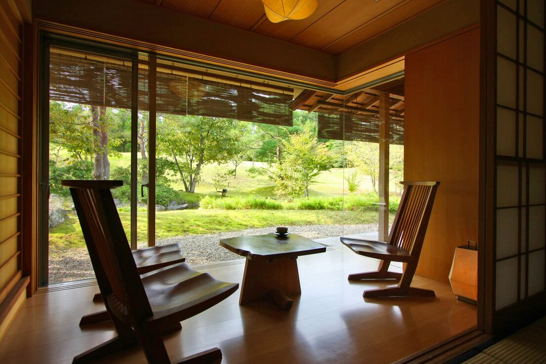 Quarto com mesa de madeira e duas cadeiras, janelas amplas com vista para o verde lá fora.