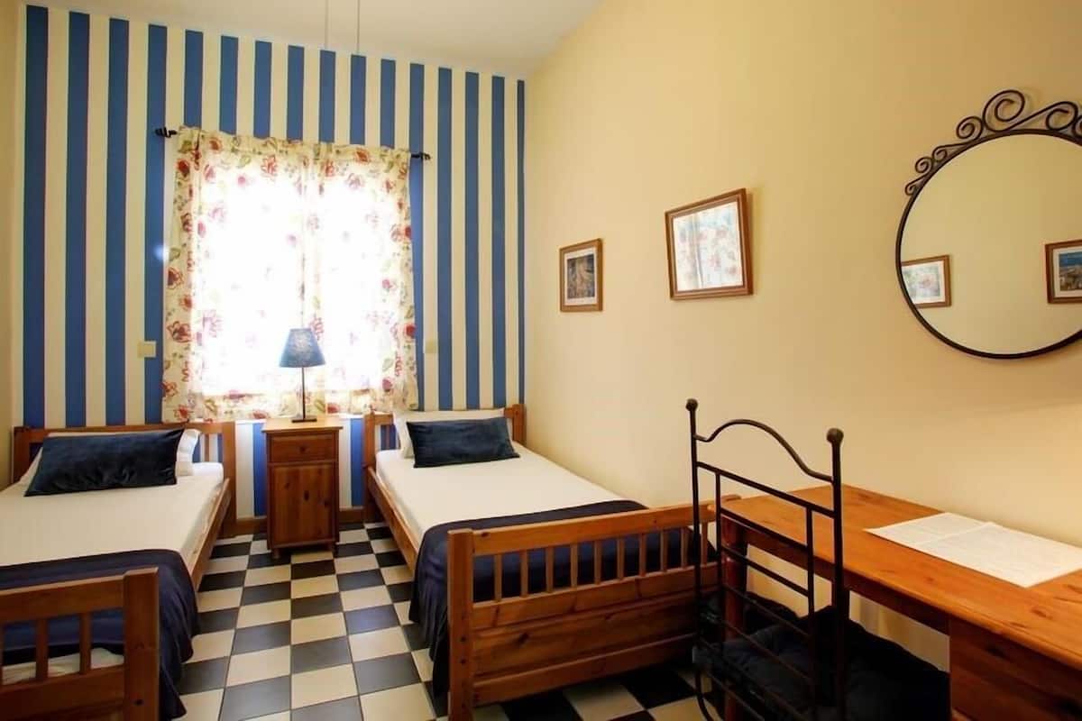 Quarto com duas camas de solteiro, uma escrivaninha de madeira, um espelho e uma janela com cortinas listradas.