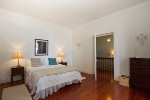 Quarto com cama, duas luminárias de cabeceira, um quadro emoldurado, cômoda e uma porta que dá para outro espaço.