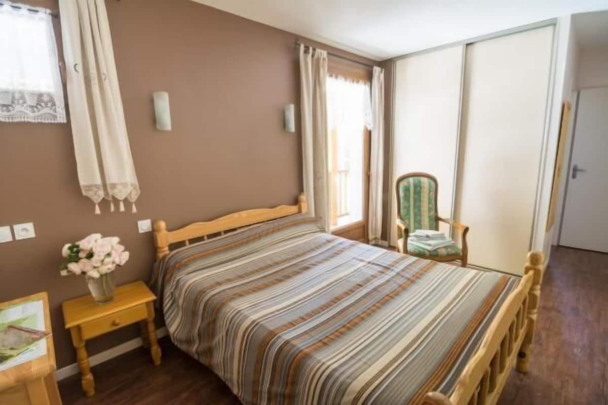 Quarto com cama de madeira, mesa de cabeceira com um vaso de flores, uma cadeira e um guarda-roupa.