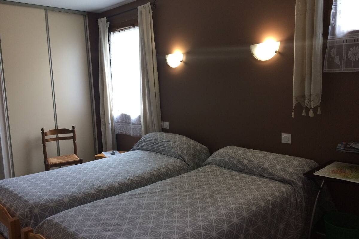 Quarto com duas camas, uma cadeira, uma mesinha e uma janela com cortinas.