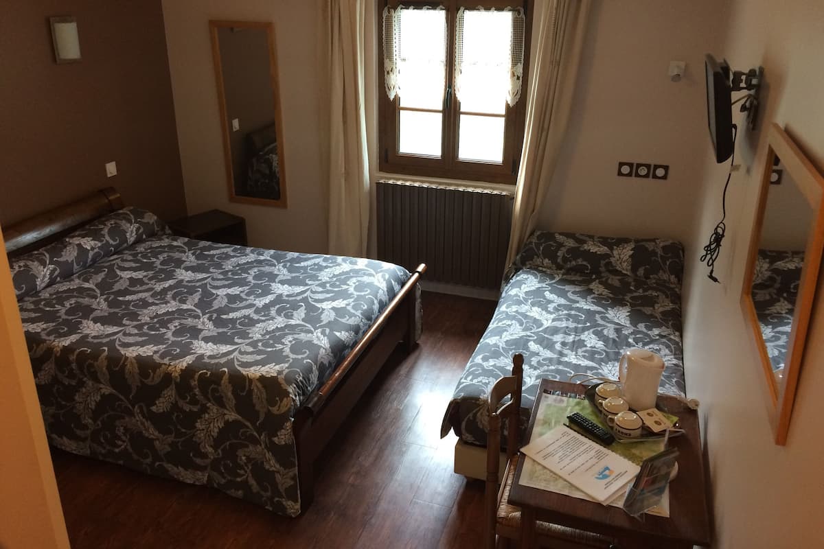 Quarto com duas camas, uma mesa de madeira com uma cadeira, um radiador e uma janela com cortinas.