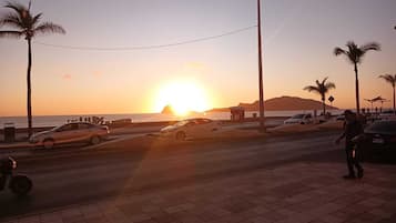 Vista do pôr do sol com palmeiras e carros estacionados ao longo de uma estrada costeira.