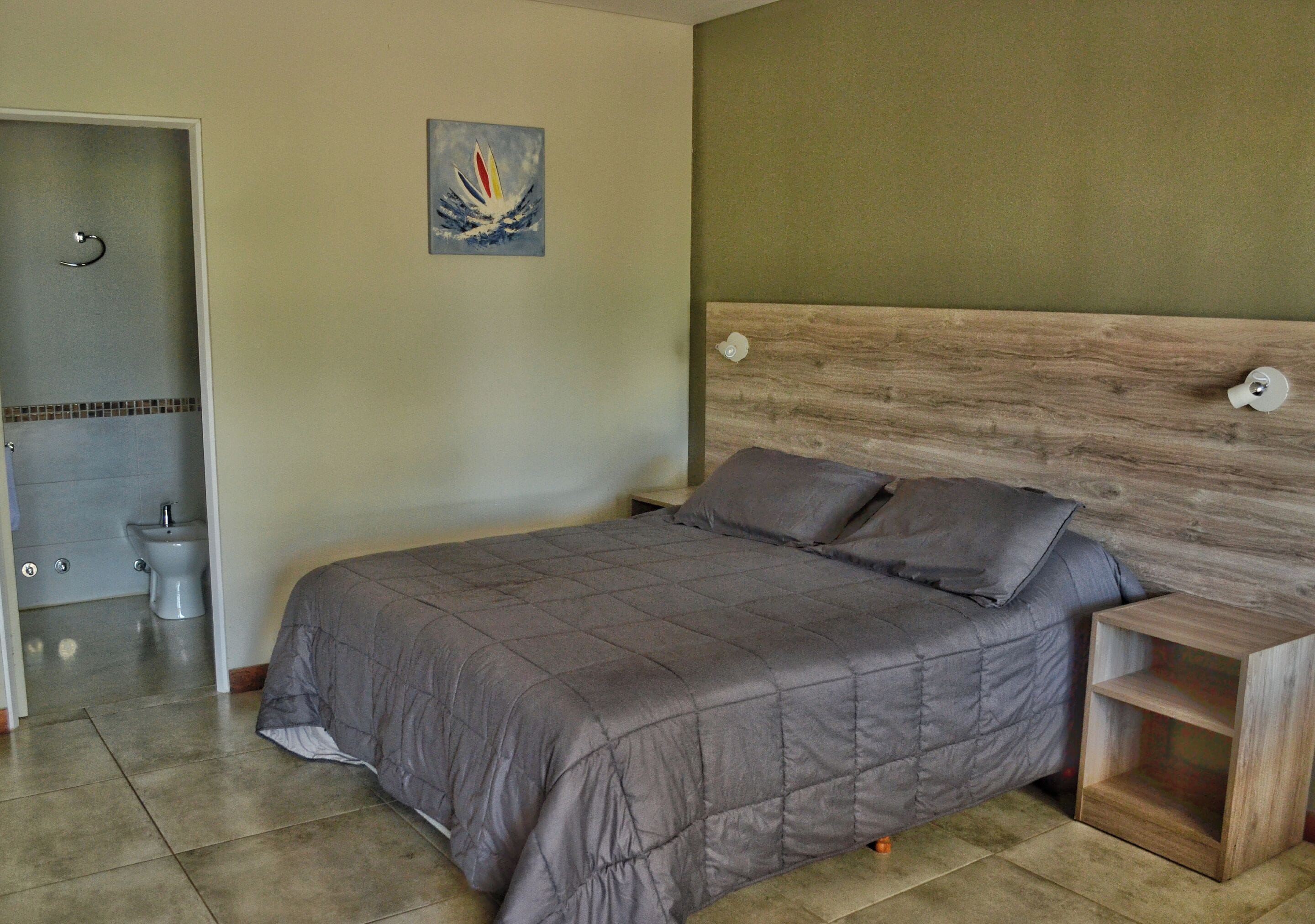 Quarto com cama, cabeceira de madeira, mesa de cabeceira, um quadro e um banheiro visível por uma porta aberta.