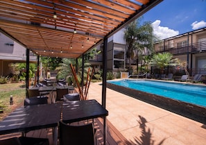 Uma área externa coberta com mesas, cadeiras, piscina e um edifício com varandas.