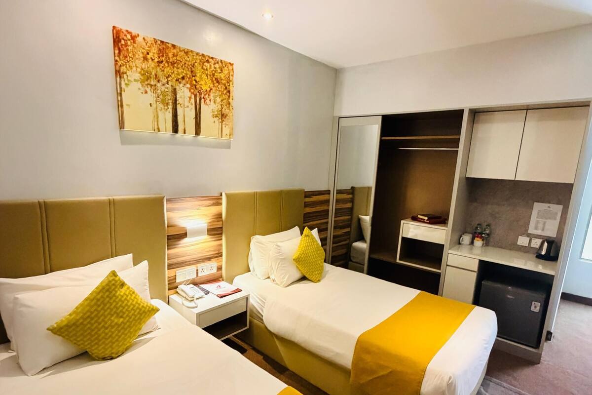 Quarto de hotel com duas camas, um quadro na parede, uma escrivaninha com cadeira e um guarda-roupa.