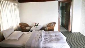 Quarto com uma cama, duas cadeiras de vime, uma mesinha com um vaso e vista para um banheiro.