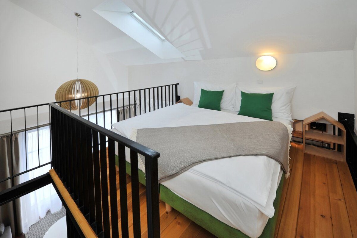 Quarto com claraboia, cama com roupa de cama verde e branca, luminária pendente e uma pequena estrutura de madeira.