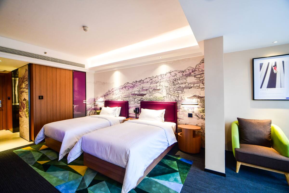 Quarto de hotel com duas camas, um mural grande na parede e uma cadeira moderna.