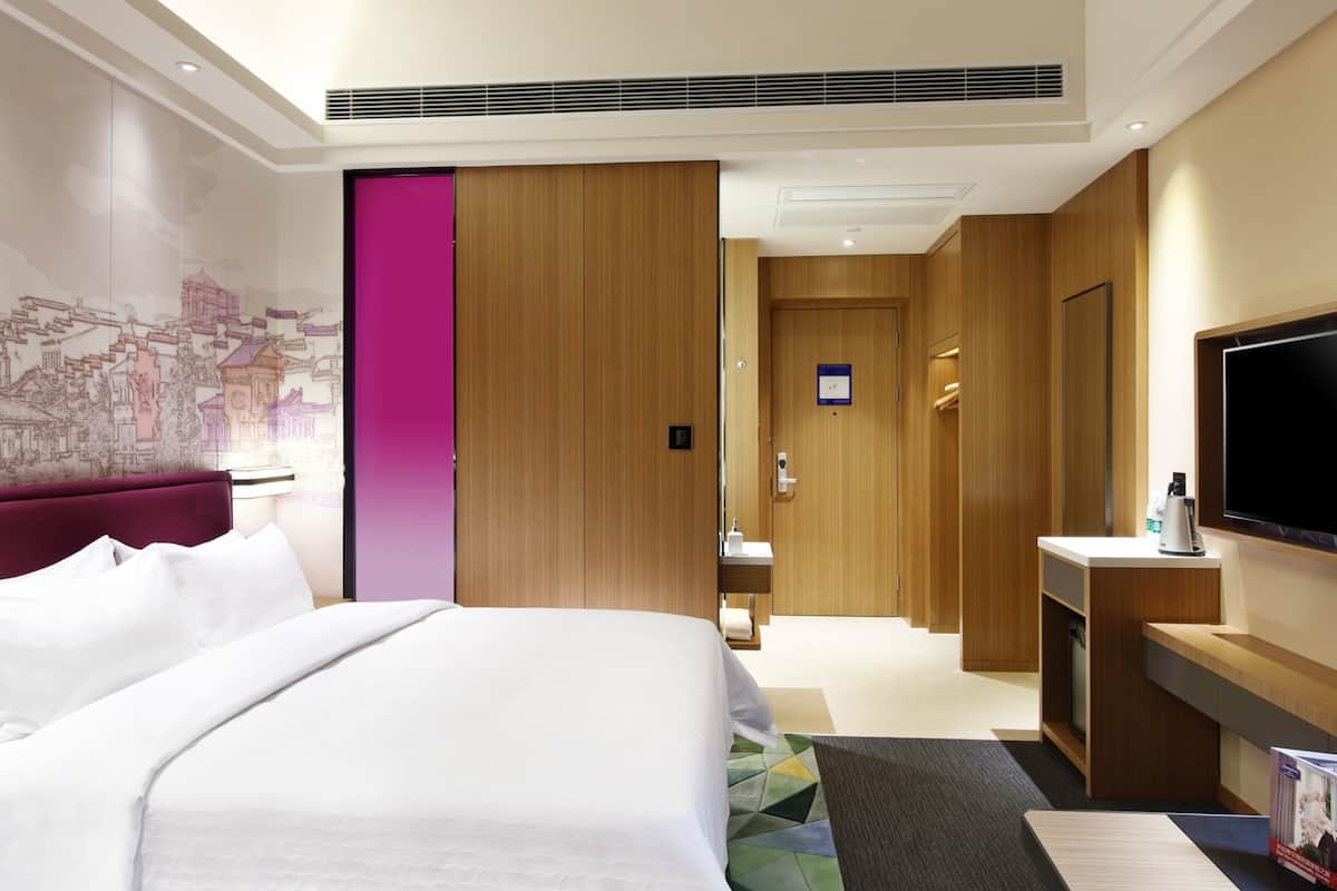 Quarto de hotel com uma cama grande, uma escrivaninha com televisão de tela plana, um banheiro com espelho e uma parede com um mural da paisagem urbana.