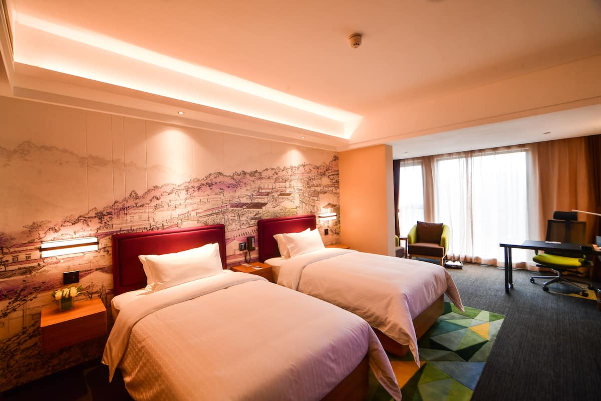 Quarto de hotel com duas camas, uma mesa de trabalho, uma cadeira e uma janela grande com cortinas.