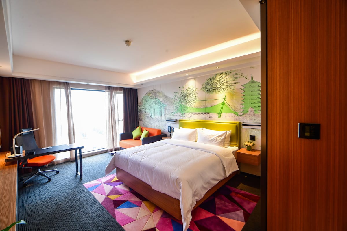 Quarto de hotel com uma cama, uma escrivaninha e uma cadeira. Há uma janela com cortinas, uma parede com um mural e uma mesa de cabeceira com um abajur.