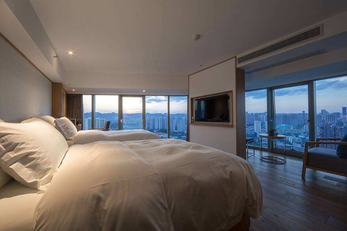 Um quarto espaçoso com uma cama grande, televisão na parede e vista para a cidade através de amplas janelas.